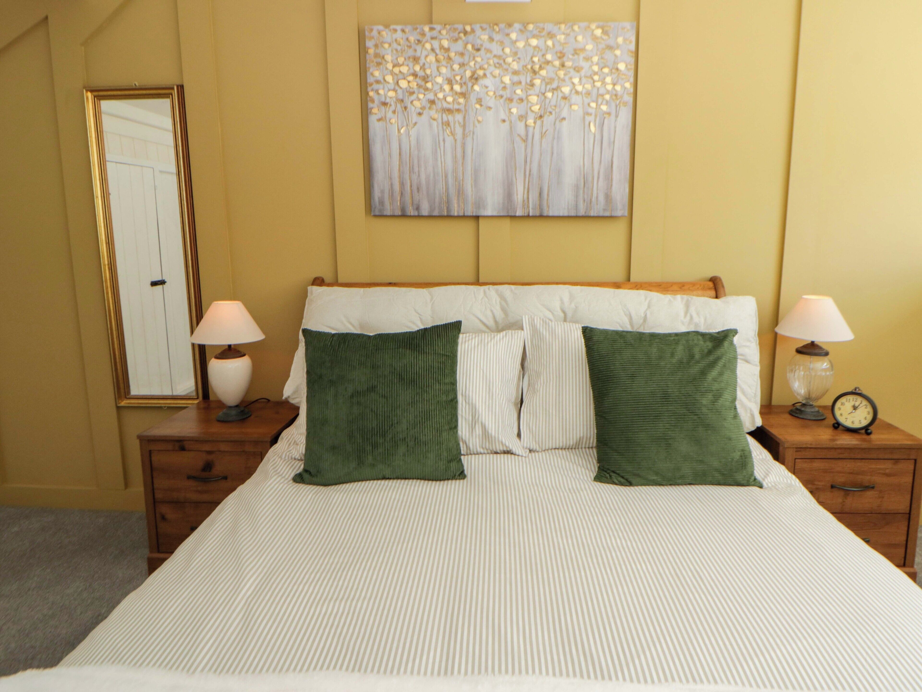 Um quarto com cama, duas mesas de cabeceira, um espelho, um quadro e um relógio.