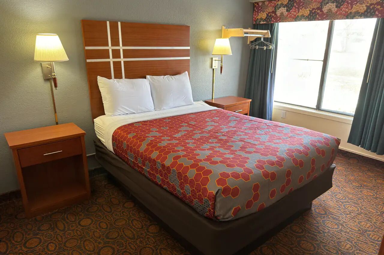 Quarto de hotel com uma cama grande, duas mesas de cabeceira, duas luminárias de parede, uma janela com cortinas e um tapete estampado.