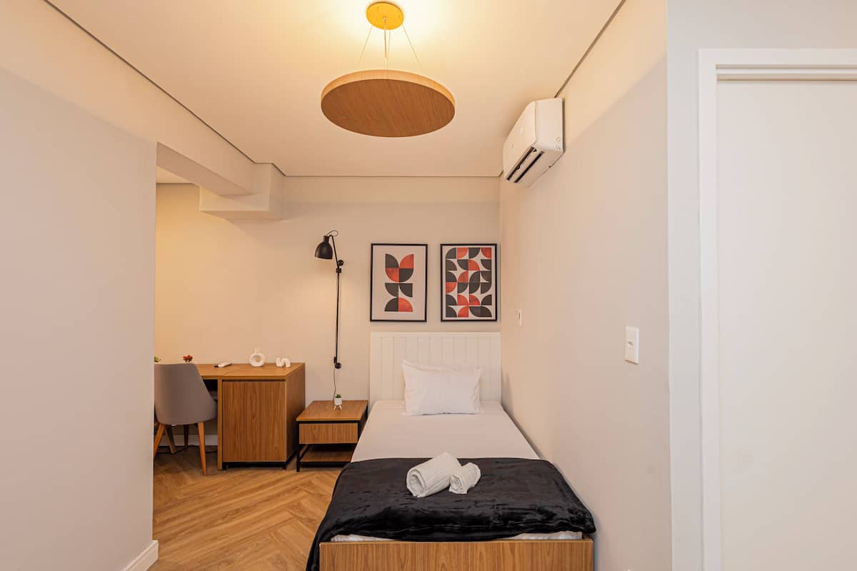 Quarto de hotel compacto com uma cama, uma escrivaninha, uma cadeira, um abajur e duas obras de arte abstratas nas paredes.