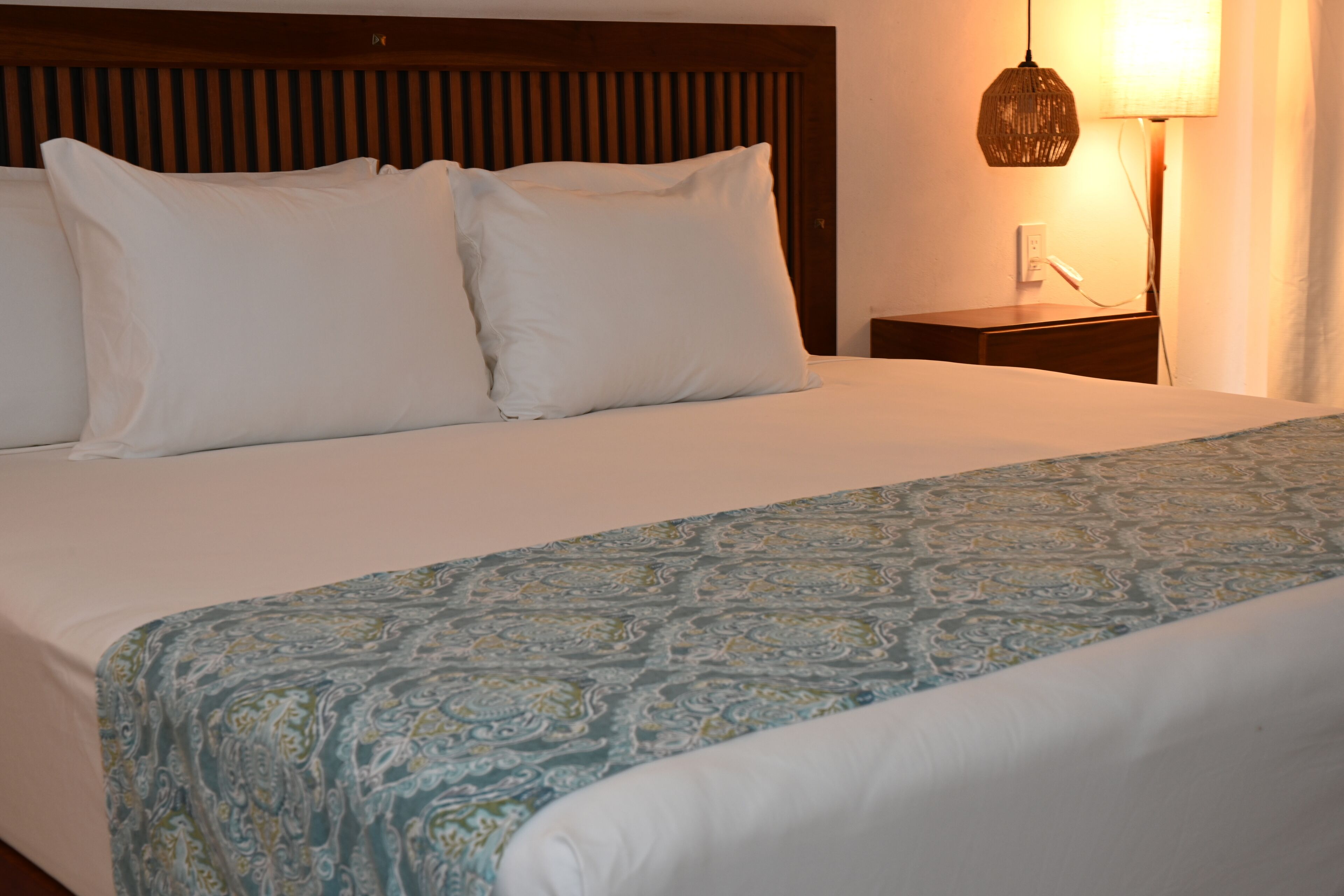 Cama bem arrumada com colcha estampada, cabeceira de madeira e abajur.