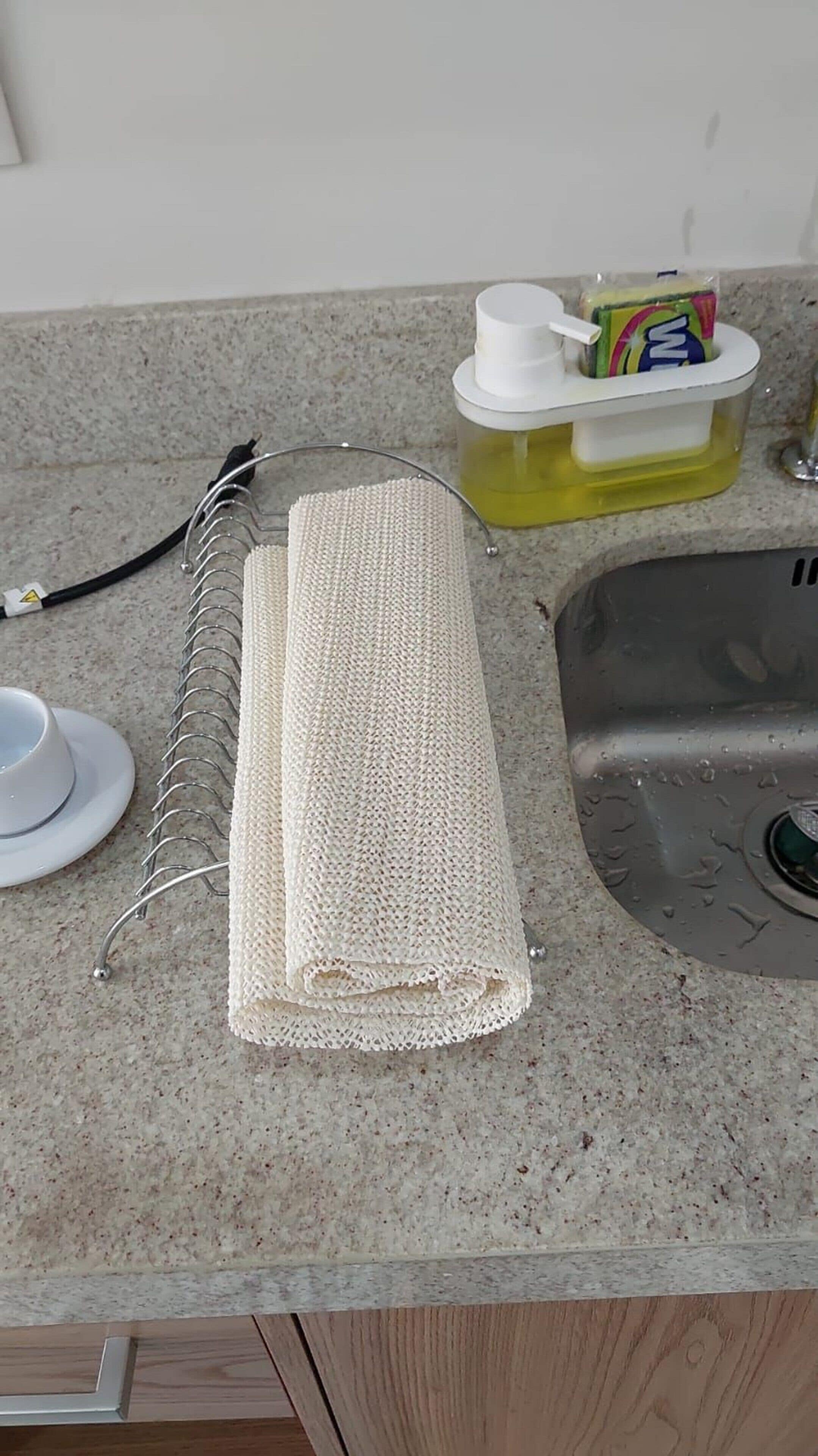 Uma bancada de cozinha com um pano branco, escorredor de louça, dosador de detergente e pia.