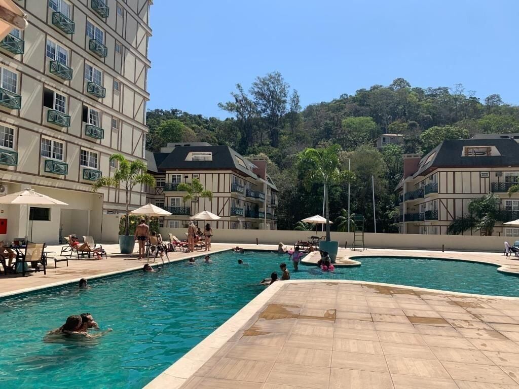 Uma área de piscina com espreguiçadeiras e guarda-sóis, cercada por edifícios com varandas.
