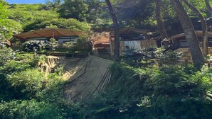 Uma cabana isolada rodeada por floresta densa, com área de estar coberta ao ar livre e escadaria que leva à entrada.