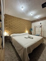 Quarto com parede em tijolos, cama com colcha branca, mesa de cabeceira com abajur e ventilador de teto.