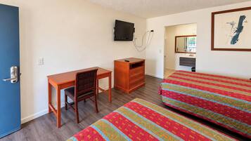Quarto com escrivaninha, cômoda, cama com colcha colorida, televisão fixada na parede e uma porta.