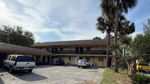 Um motel com um edifício de dois andares, estacionamento e uma pequena área de jardim com palmeiras.