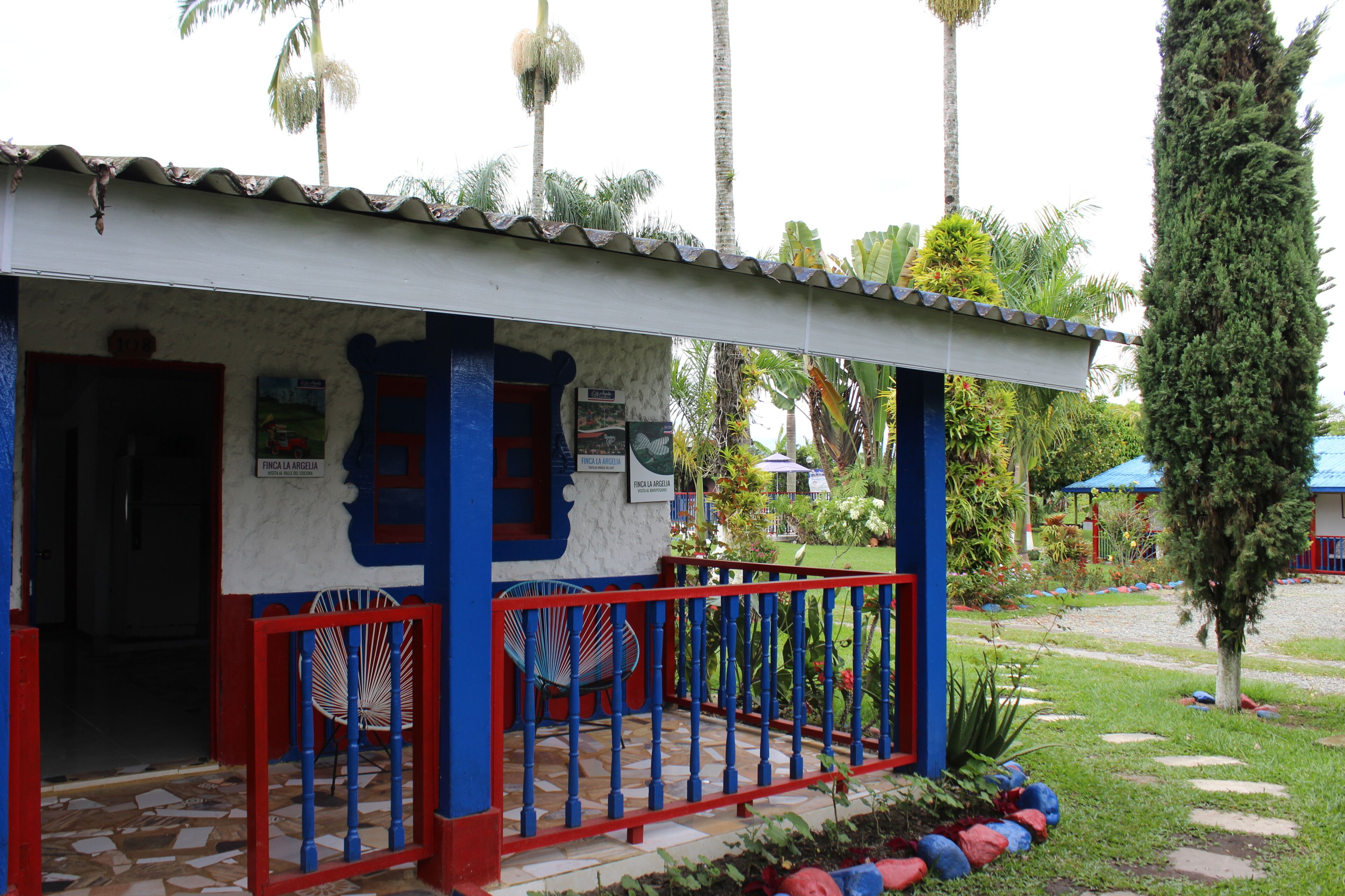 Um edifício com entrada em azulejos, guarda-corpo vermelho e azul, e um jardim com palmeiras e um caminho.