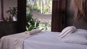 Uma cama com roupa branca, uma rede e vista para o verde lá fora.