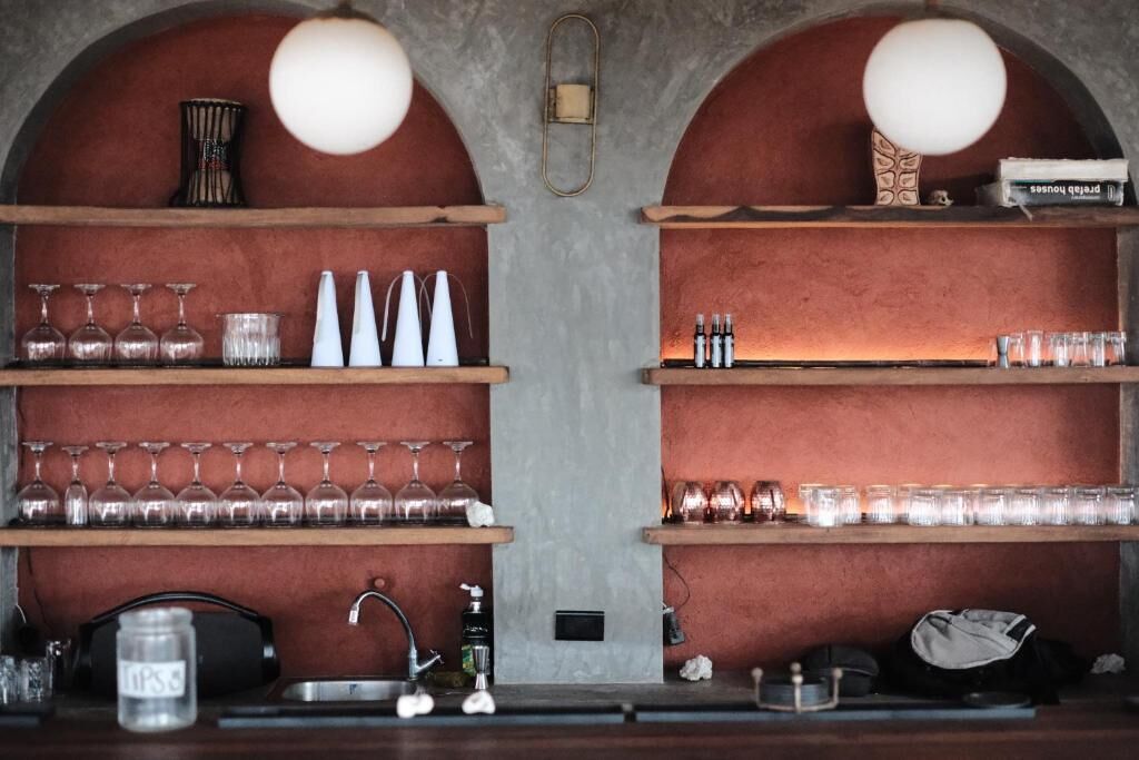 Uma área de bar com prateleiras de copos e garrafas, uma pia e uma bolsa no balcão.