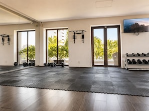 Uma academia com equipamentos de ginástica, TV e amplas janelas com vista para palmeiras.