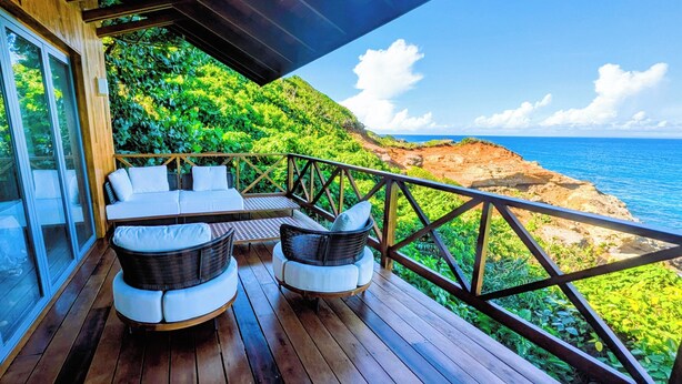 Um deck de madeira com mesa e cadeiras, com vista para uma paisagem costeira com vegetação e um céu azul e límpido.