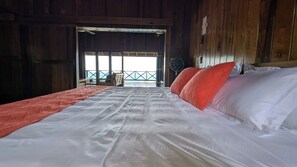 Uma cama com roupa de cama branca e laranja num quarto de madeira com vista para o mar.