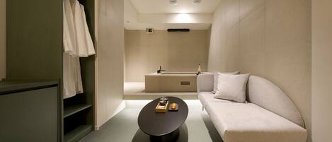 Quarto de hotel moderno com sofá, uma pequena mesa com uma garrafa e uma caixa, banheira e guarda-roupa.