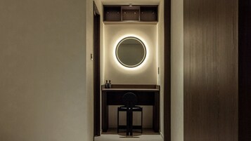 Corredor estreito que leva a uma toucador com espelho redondo, uma pequena mesa e um prateleira com garrafas.