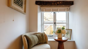 Quarto de canto com uma cadeira estofada, uma pequena mesa de madeira com um vaso de flores, uma janela com persianas e um quadro na parede.