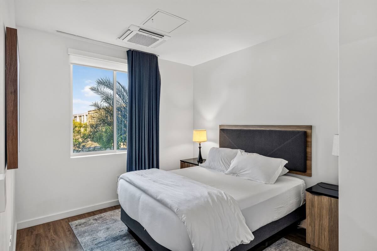 Quarto de hotel moderno com uma cama grande, uma mesinha de cabeceira, uma luminária, uma janela com cortinas e vista para palmeiras.