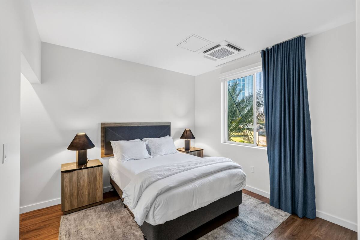 Quarto com cama, duas mesas de cabeceira, janela com cortinas e um respiradouro no teto.