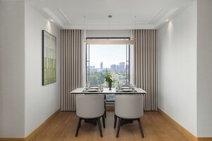 Uma área de refeições com uma mesa posta para dois, vista da cidade através de uma janela ampla e uma obra de arte emoldurada na parede.