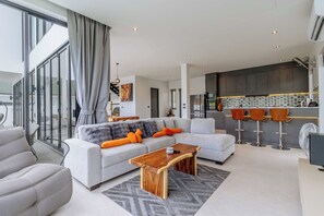 Uma sala de estar moderna com um sofá cinzento, mesa de centro de madeira e uma área de cozinha com armários escuros e painel de azulejos.