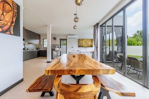 Interior moderno com mesa de jantar de madeira, cozinha e grandes janelas.