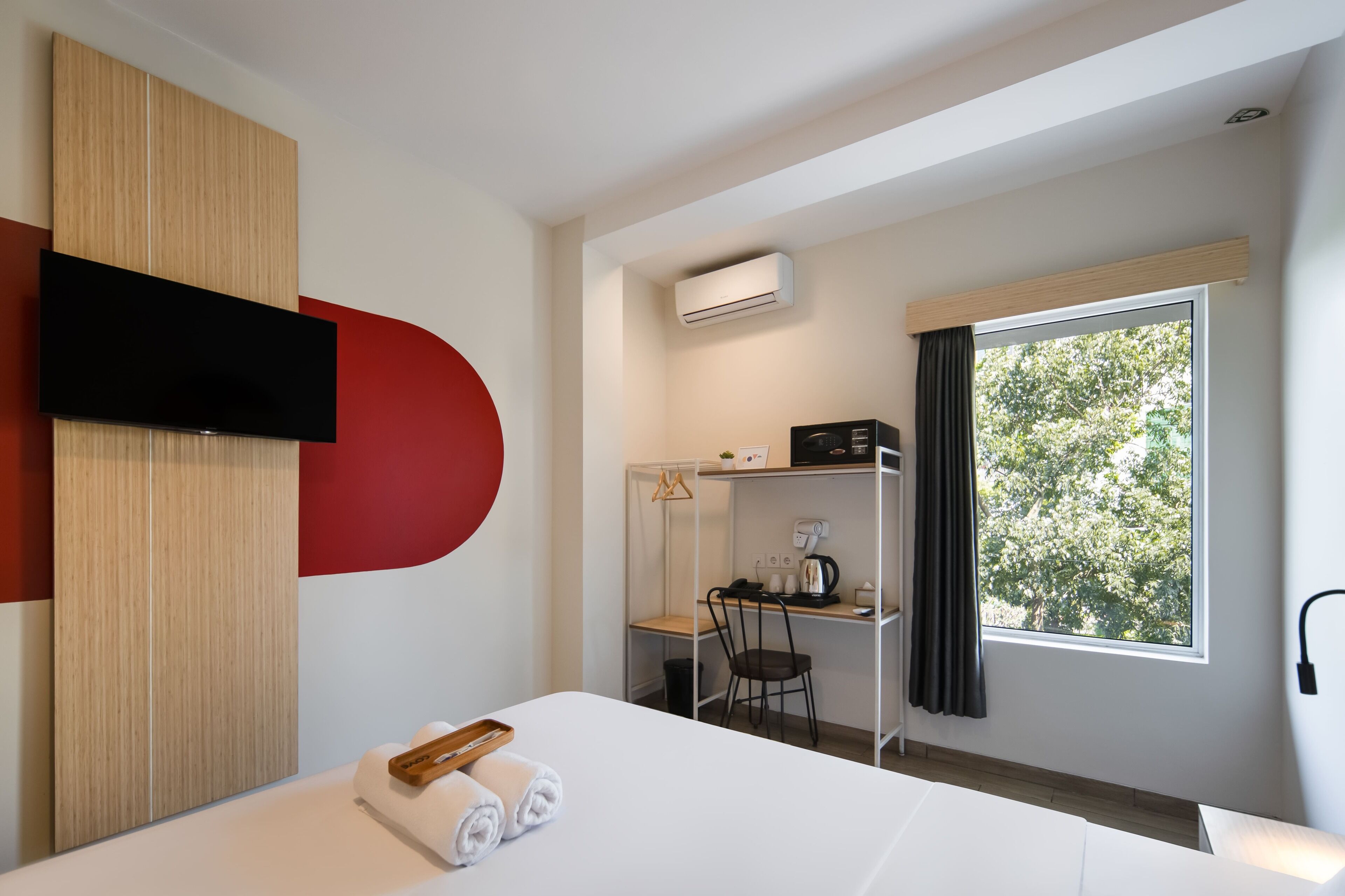 Quarto de hotel com cama, escrivaninha, cadeira, ar condicionado, janela com vista e design de parede em vermelho e branco.