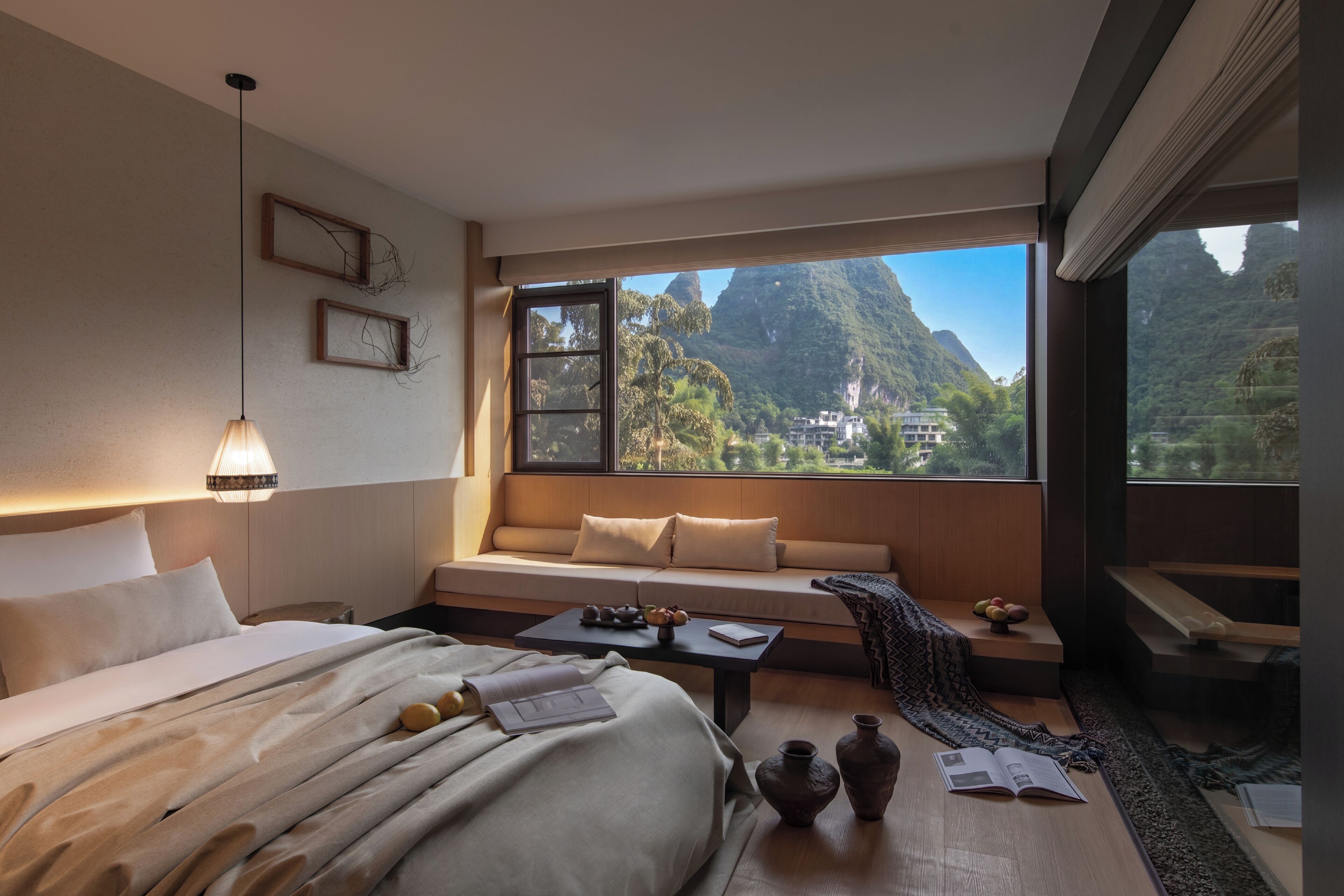Quarto de hotel moderno com uma cama grande, área de estar com vista para montanhas, uma mesinha com livros e um vaso, e uma janela ampla.
