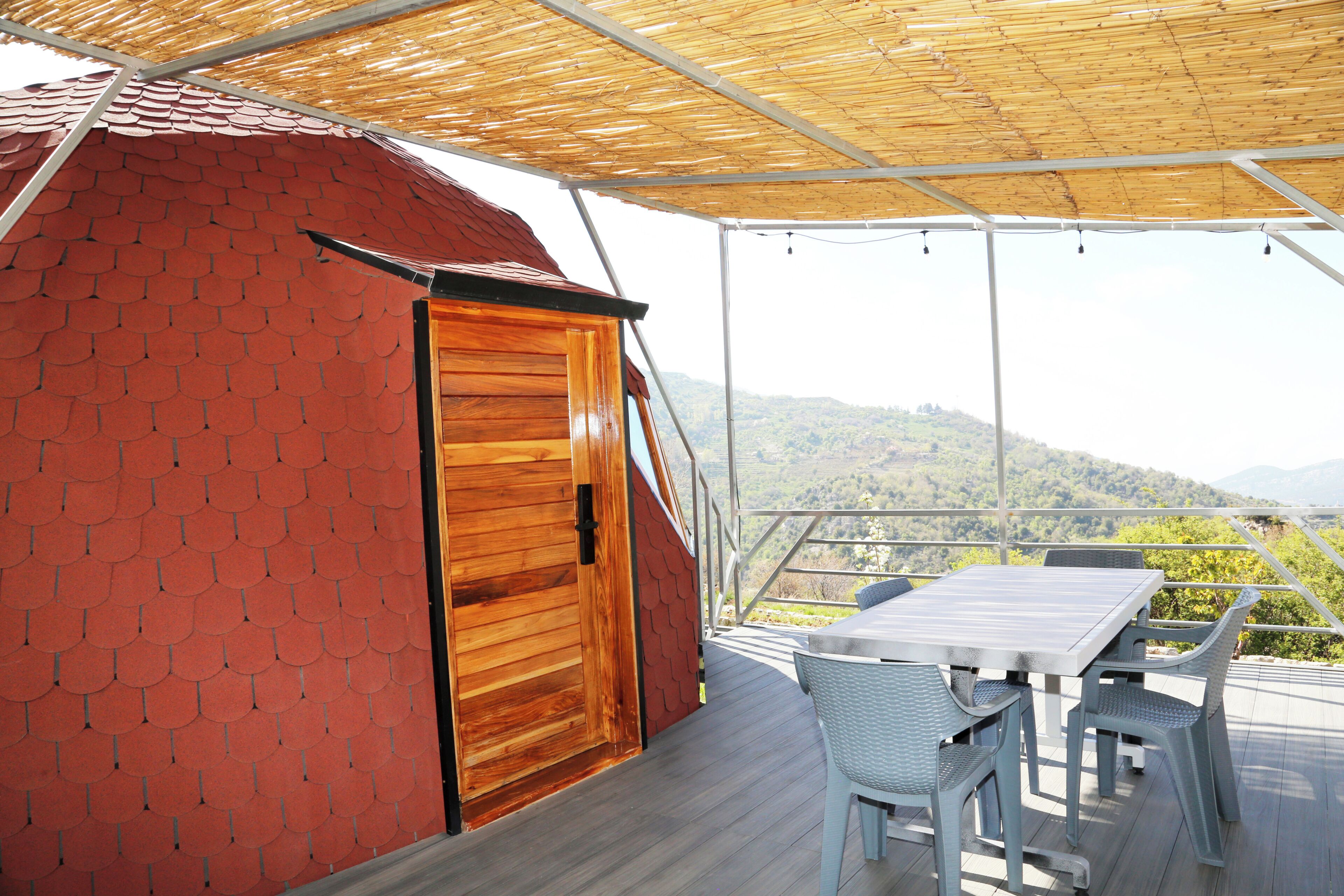 Uma cabana de madeira com exterior vermelho, uma porta deslizante e uma mesa com cadeiras num terraço elevado com vista para a paisagem.