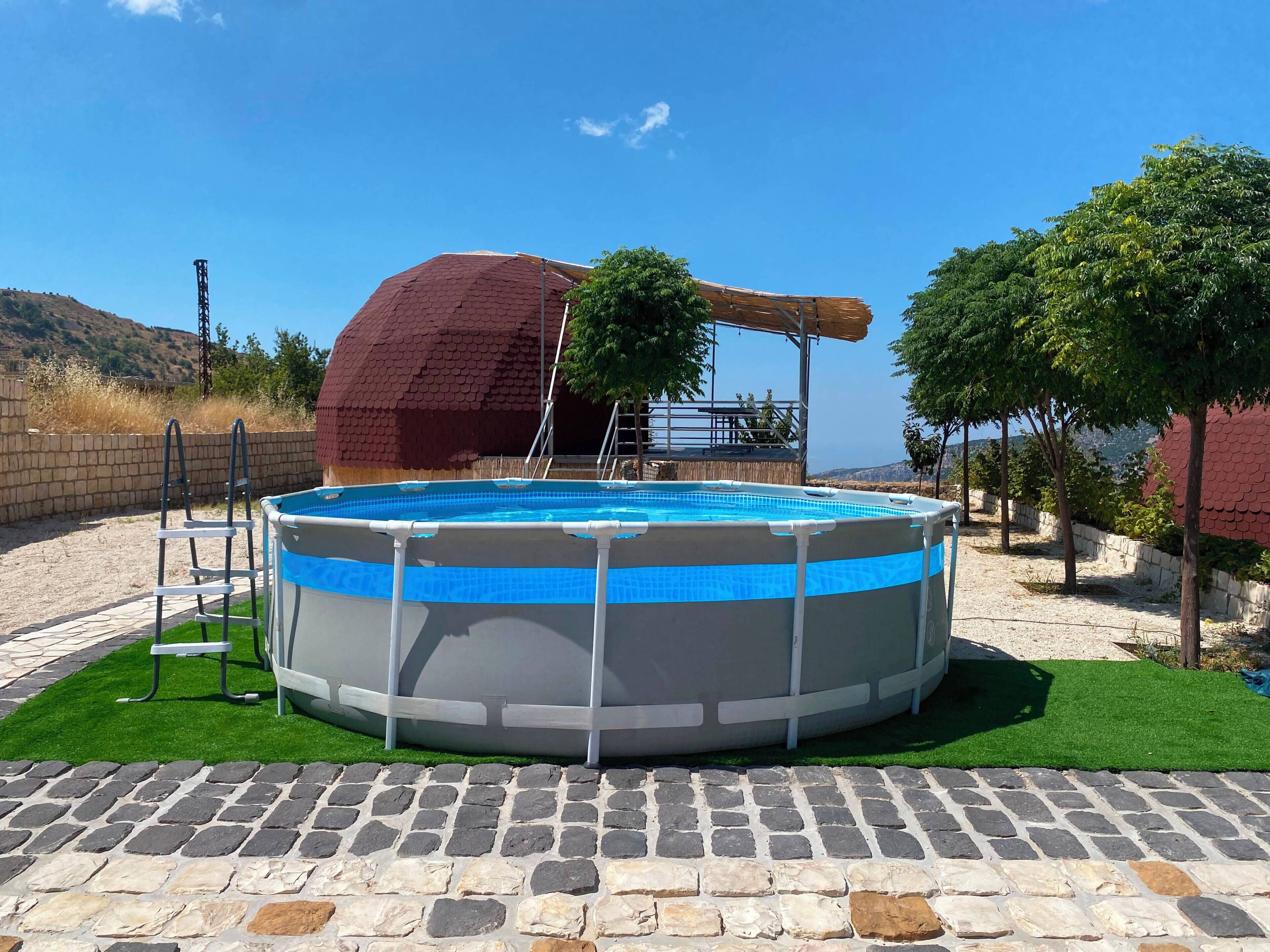 Uma piscina redonda com escada, rodeada por um pátio de pedra e um gramado.