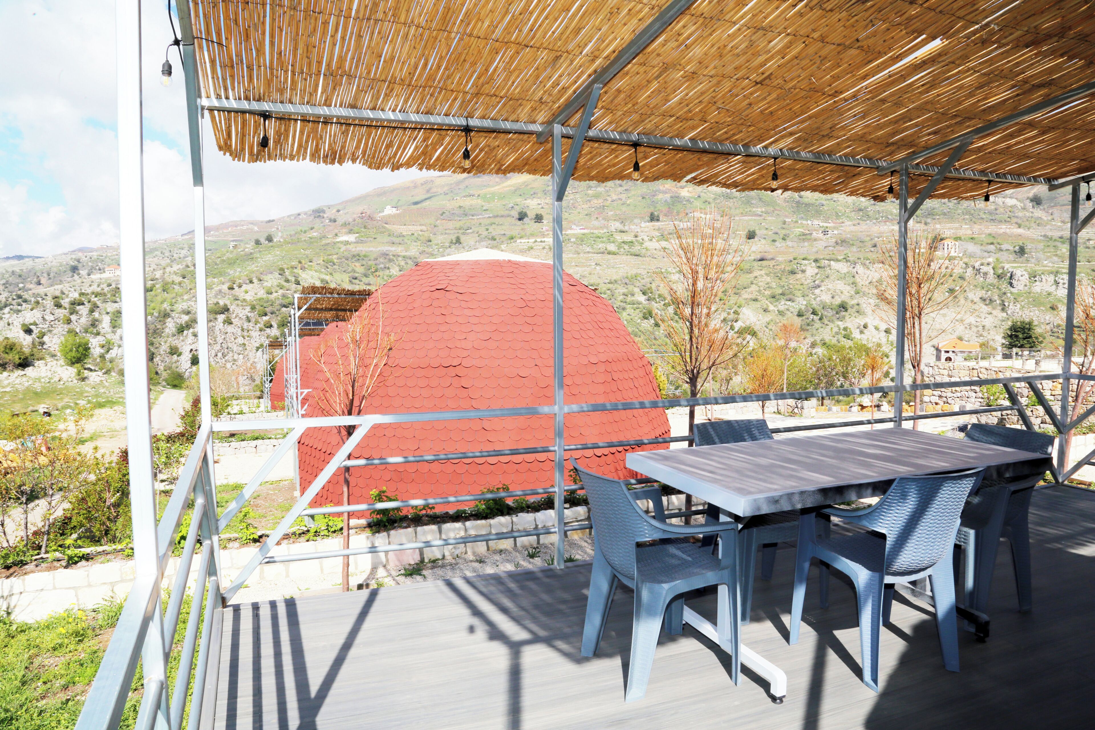 Uma área de estar coberta ao ar livre, com mesa e cadeiras, com vista para um edifício de telhado vermelho e uma paisagem montanhosa.