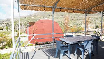 Uma área de estar coberta ao ar livre, com mesa e cadeiras, com vista para um edifício de telhado vermelho e uma paisagem montanhosa.