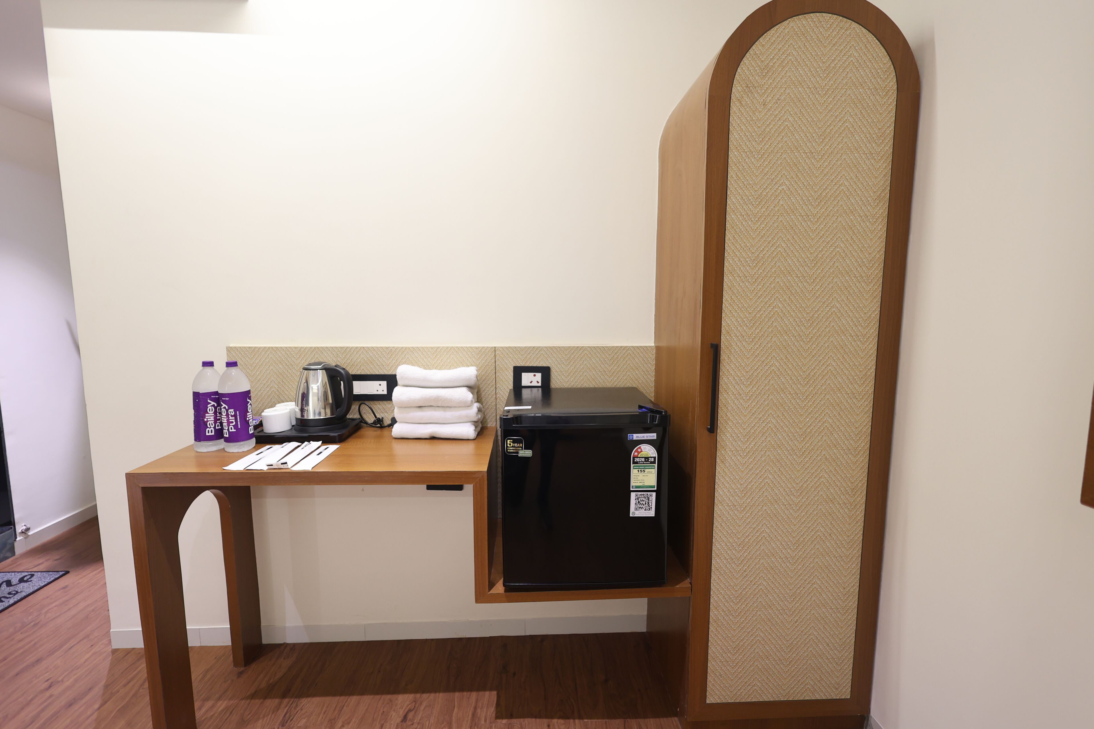 Quarto de hotel com escrivaninha de madeira, frigorífico preto, chaleira, pilha de toalhas brancas e duas garrafas de água Bailey's Pure Pura.