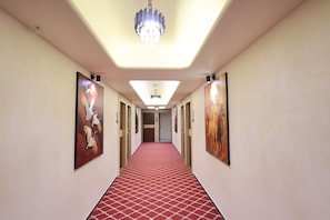 Um corredor de hotel com carpete estampado em vermelho, um candelabro e obras de arte nas paredes.