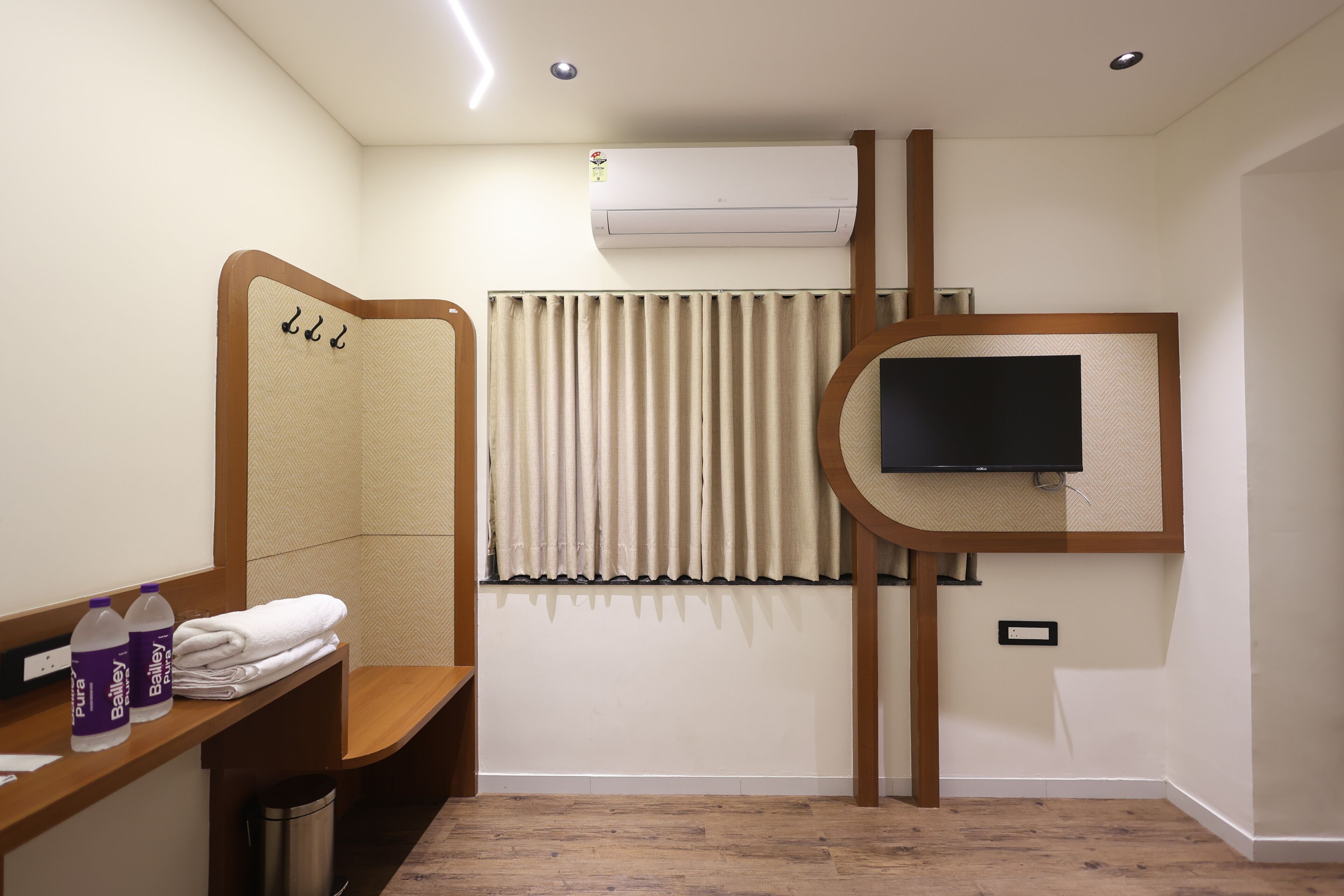 Quarto de hotel com um banco de madeira, uma televisão fixada na parede, um aparelho de ar condicionado e uma janela com cortinas.