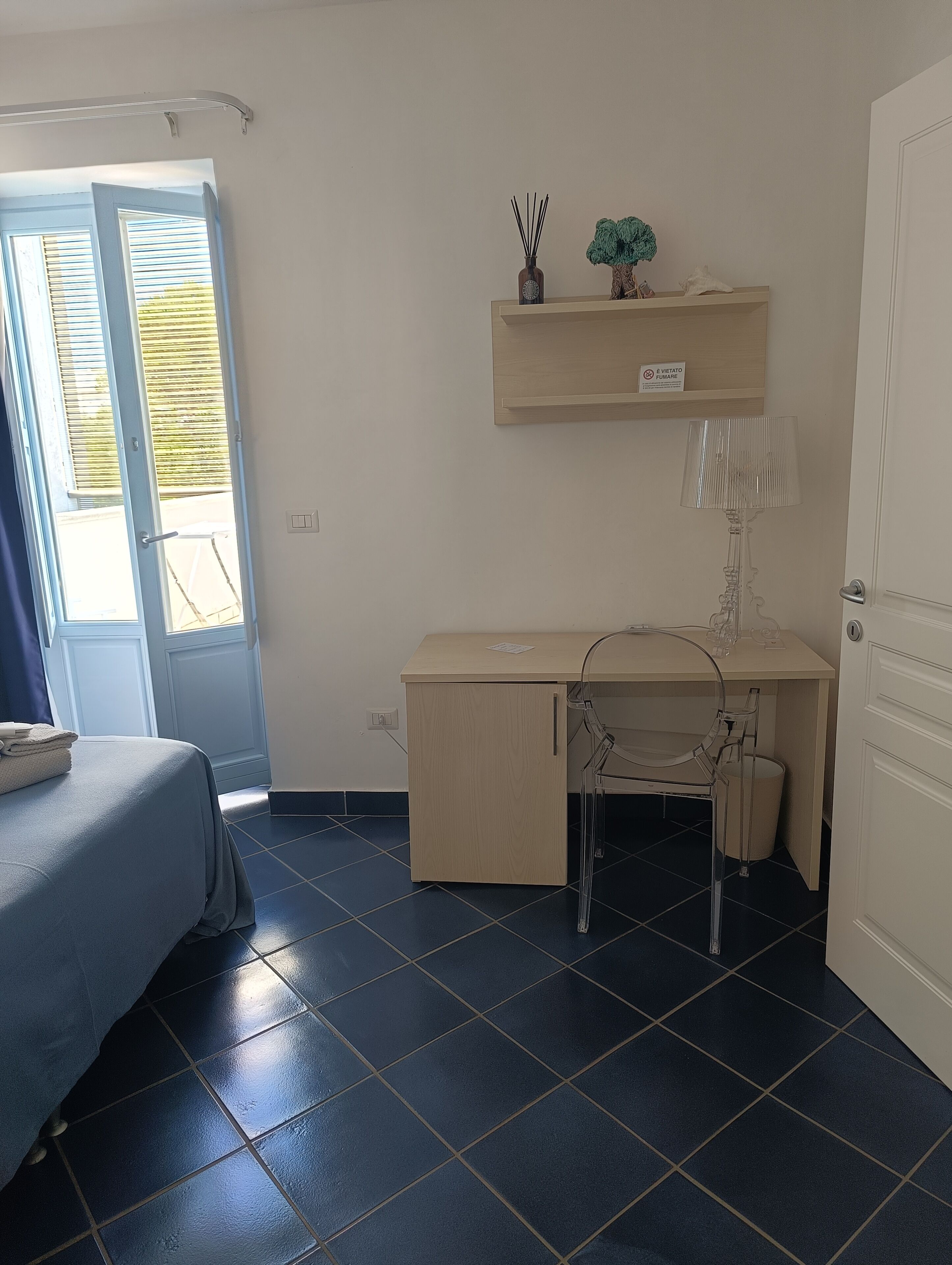Quarto com uma cama, uma escrivaninha, uma cadeira, uma estante com objetos decorativos e uma porta.