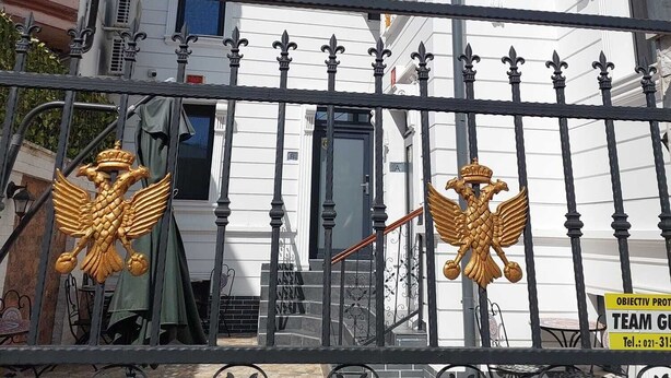 Um edifício com fachada branca, telhado preto e cerca de metal negra decorada com águias douradas.