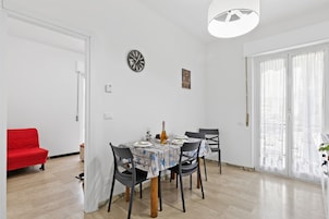 Uma sala de jantar com uma mesa posta para quatro pessoas, um sofá vermelho, um relógio e uma janela com cortinas.