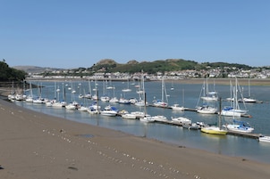 Uma marina com vários barcos atracados na costa, uma praia de areia e a paisagem de uma cidade costeira com casas e colinas ao fundo.