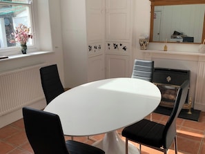 Uma sala de jantar com uma mesa branca redonda, cadeiras pretas, um vaso com flores, um radiador e um espelho na parede.