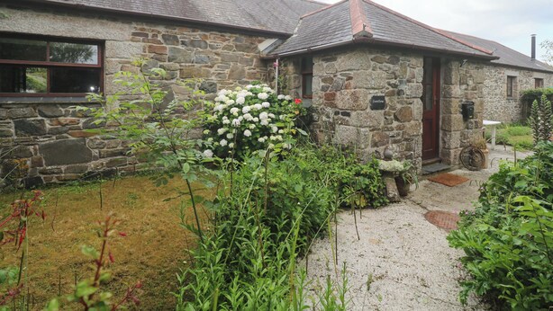 Uma casa de pedra com um caminho de cascalho que leva à entrada, rodeada por vegetação e um pequeno jardim.