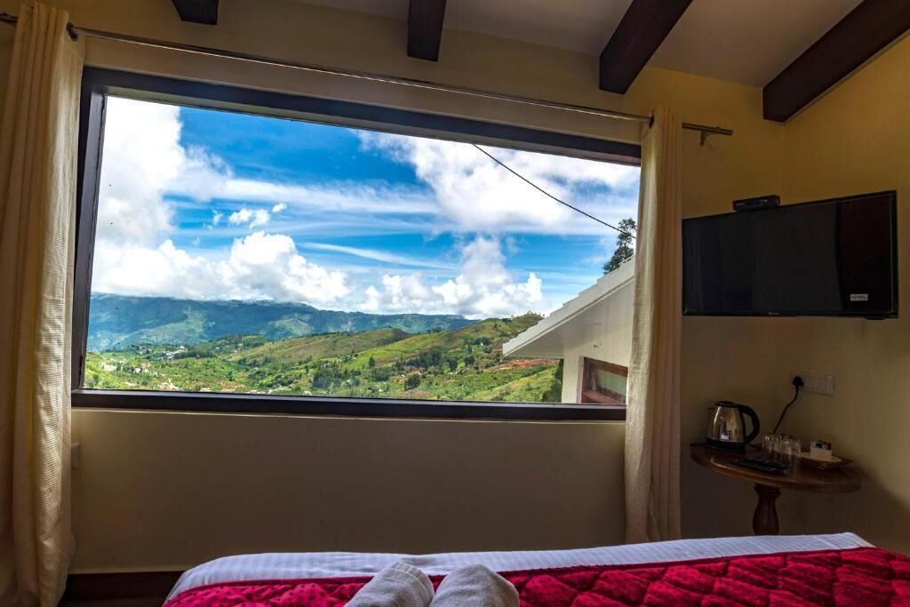 Quarto com uma janela grande, uma cama, uma mesinha com chaleira e televisão na parede.