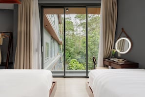 Quarto de hotel com duas camas, uma pequena escrivaninha, um espelho e vista para o verde lá fora.