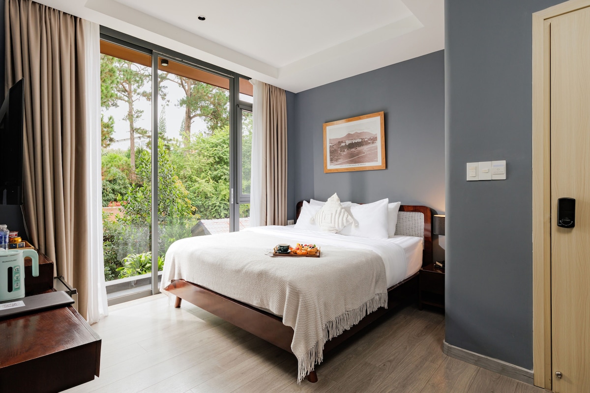 Quarto de hotel com uma janela grande, cama com roupa de cama branca, quadro emoldurado na parede e mesa de cabeceira de madeira.
