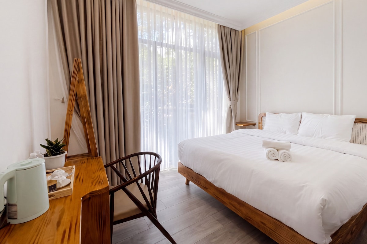 Quarto de hotel com uma cama, uma escrivaninha de madeira, uma cadeira, uma cafeteira e uma janela com cortinas.