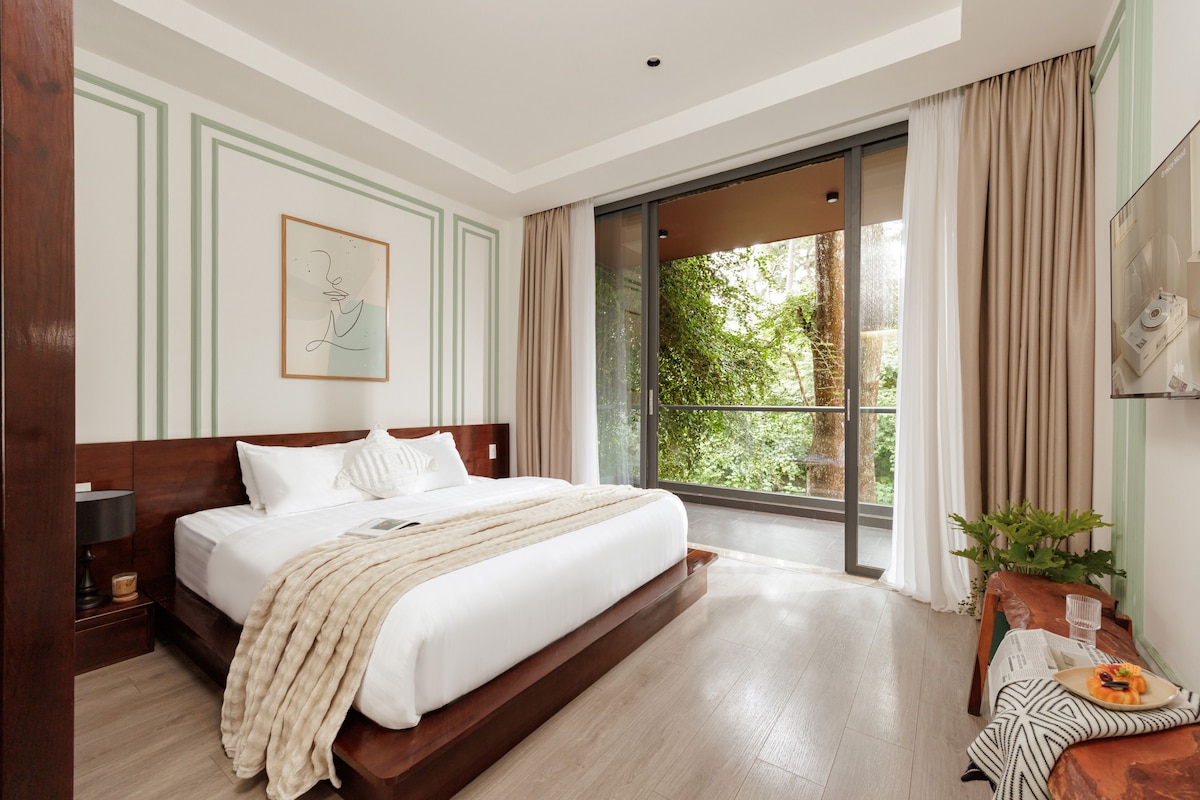 Quarto com cama grande, mesa de cabeceira, cortinas e vista para o verde lá fora.