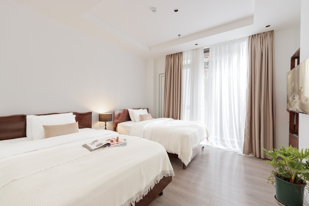 Quarto de hotel com duas camas, mesa de cabeceira com abajur, uma janela grande com cortinas e uma planta em vaso.