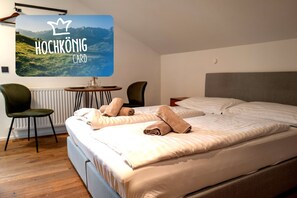 Quarto de hotel com cama, duas cadeiras, uma mesinha e uma tela na parede exibindo 'Hochkönig CARD'.