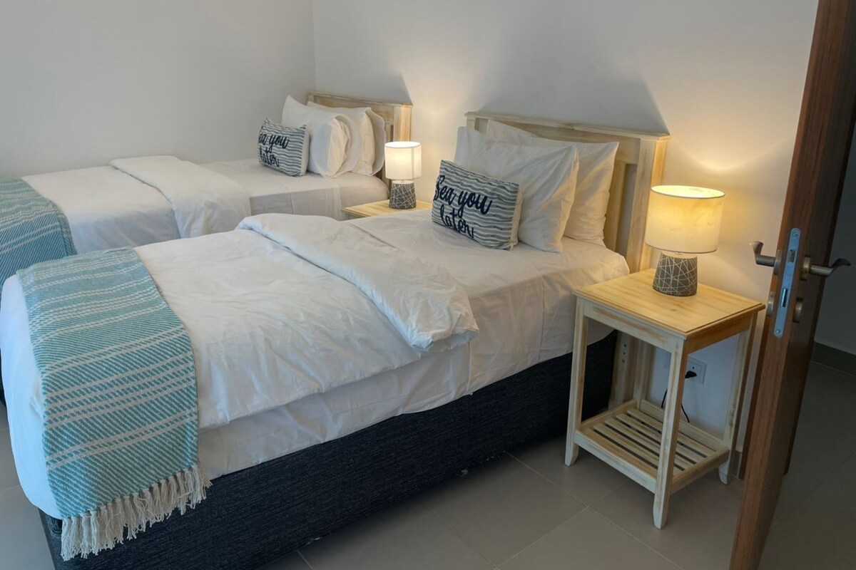 Quarto com duas camas, mesas de cabeceira com abajures, uma porta e uma parede.