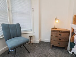Quarto com uma cadeira azul, cómoda de madeira, luminária e uma pequena mesa redonda.