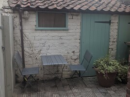 Uma pequena área externa com mesa e duas cadeiras, uma planta em vaso e uma porta verde.