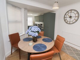 Uma área de refeições com uma mesa de madeira redonda, quatro cadeiras laranja, um vaso com flores azuis e um relógio de parede.